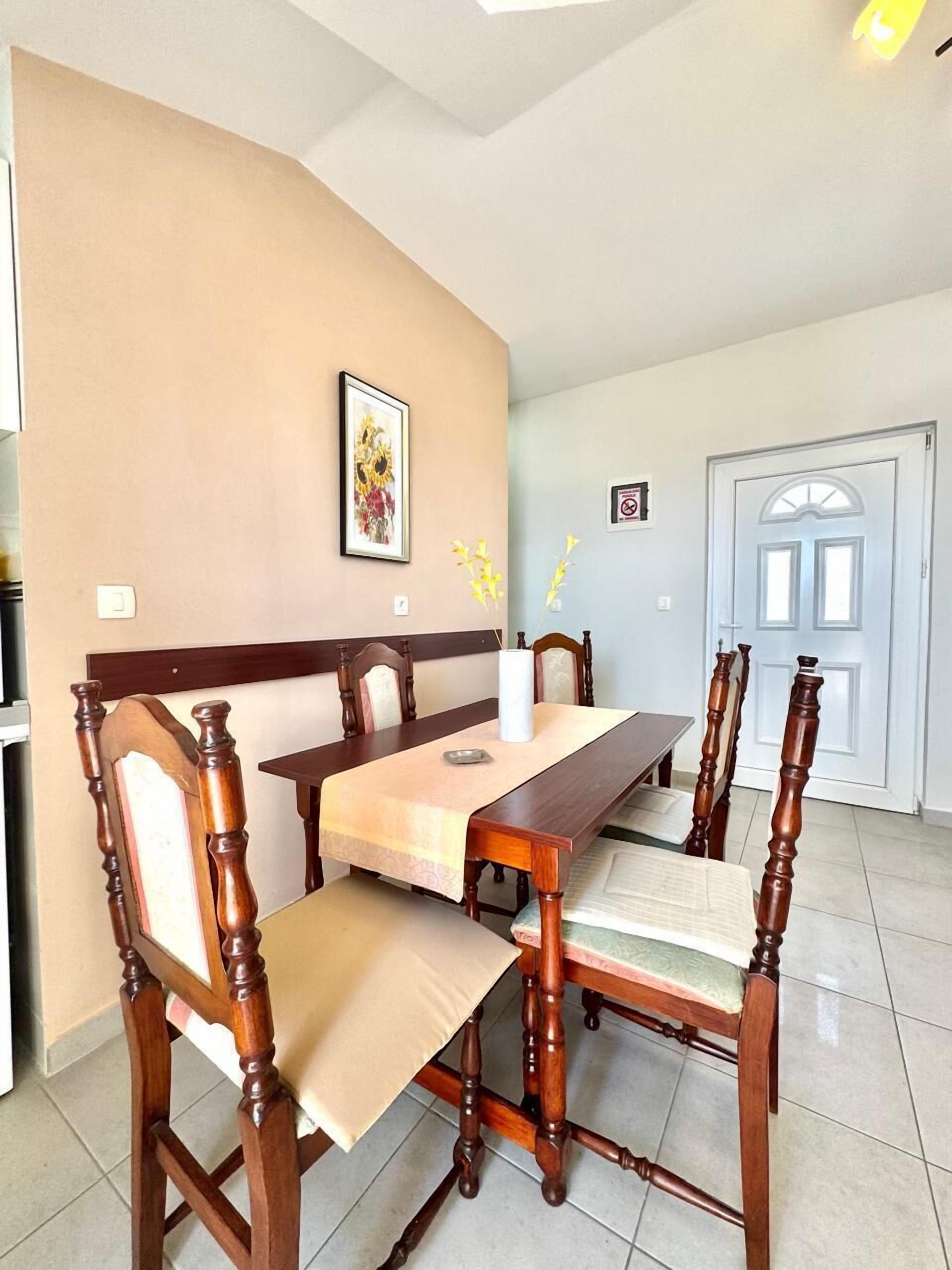 Uma área de refeições com mesa e cadeiras de madeira, um quadro na parede e uma porta ao fundo.