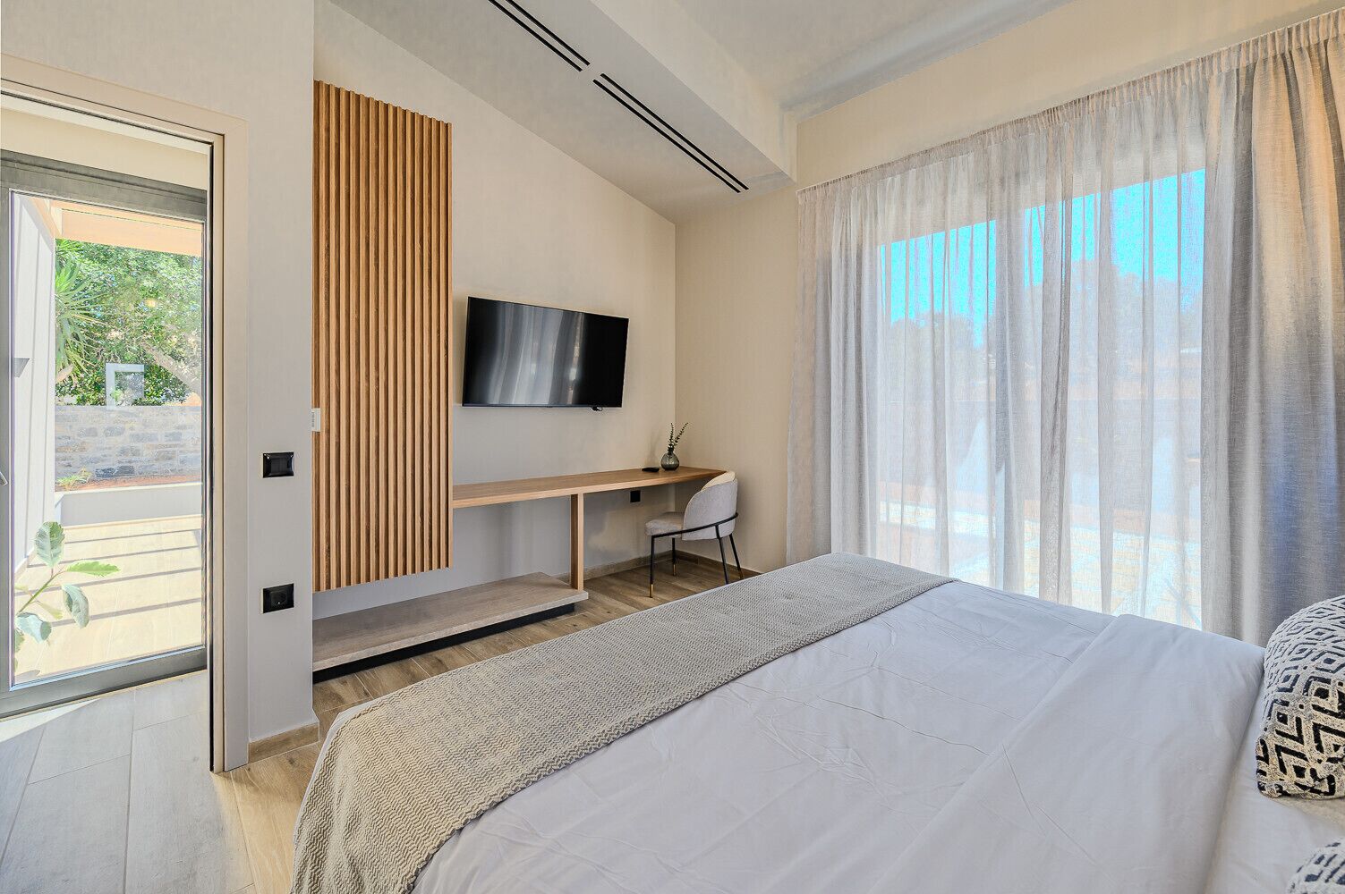Quarto de hotel moderno com uma cama grande, uma escrivaninha com cadeira, televisão de tela plana na parede e uma janela ampla com cortinas leves.