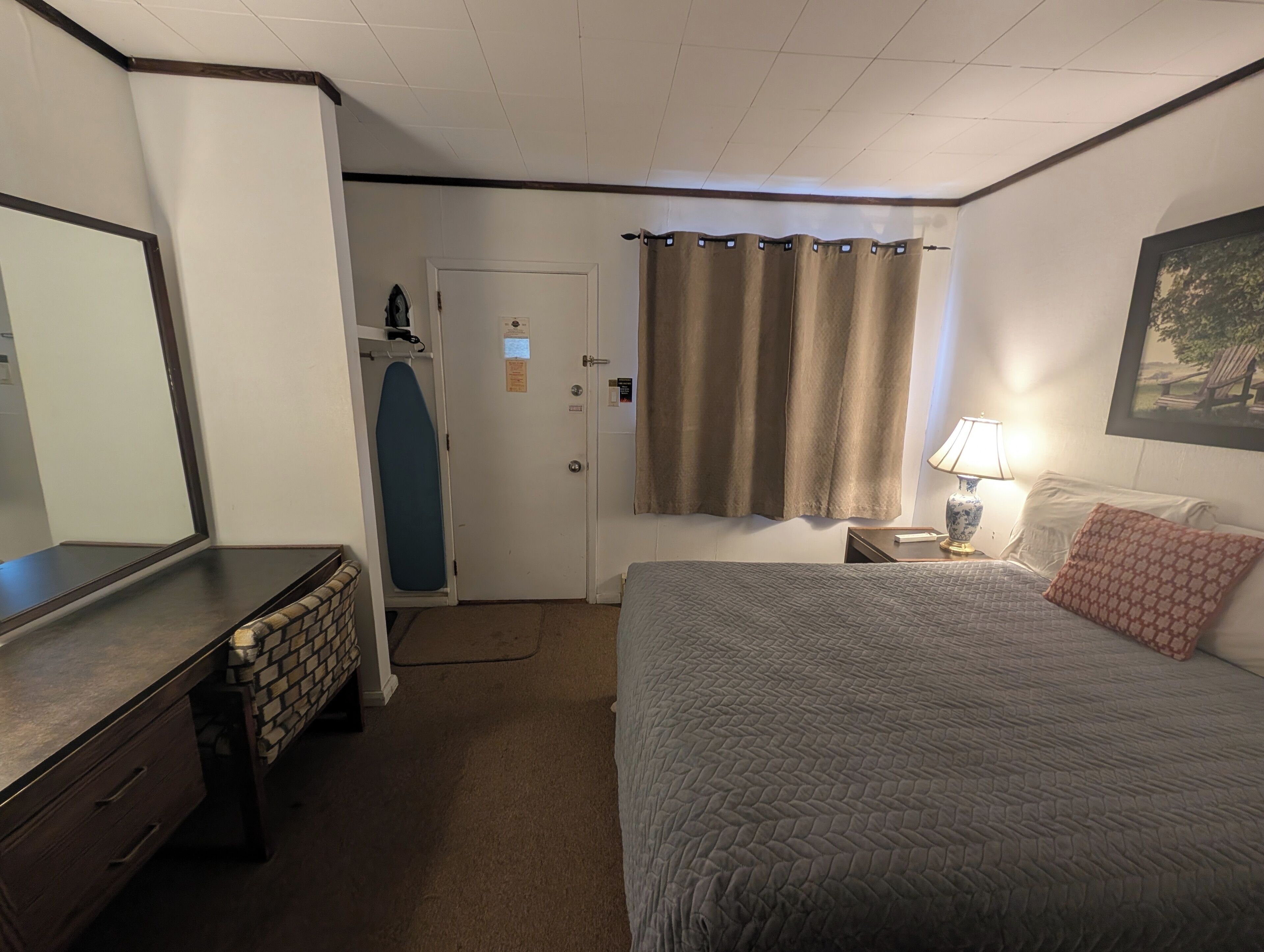 Quarto de hotel com cama, escrivaninha, espelho, porta e janela com cortinas.
