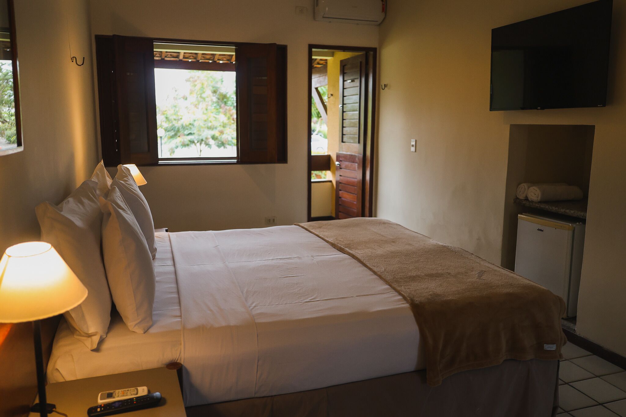 Cama bem arrumada com edredom branco e marrom, luminária de cabeceira e controle remoto em uma mesinha de cabeceira.