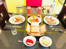 Uma mesa de jantar com diversos pratos, incluindo um prato com linguiça, um prato com salada e um prato com torradas. Há também dois copos de uma bebida vermelha e uma garrafa de água.