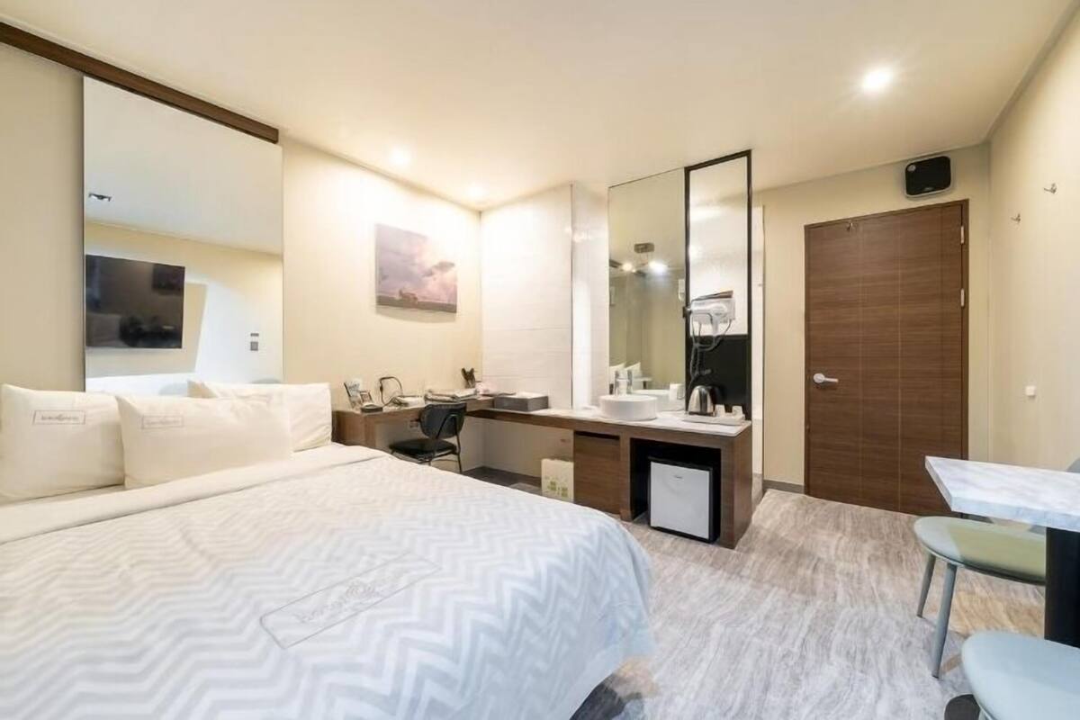 Quarto de hotel com uma cama, escrivaninha, cadeira, espelho e guarda-roupa.