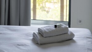 Cama bem arrumada com toalhas dobradas e um controle remoto, uma janela com cortinas e vista para o verde lá fora.