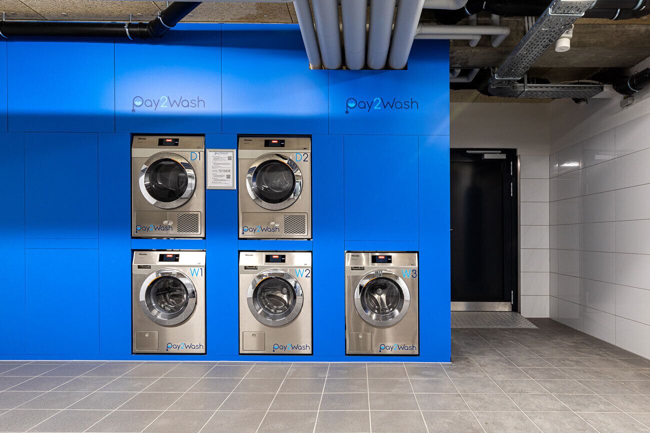 Quatro máquinas de lavandaria automática Pay2Wash alinhadas contra uma parede azul em um espaço público.