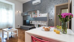 Uma sala de estar moderna com um sofá branco, uma TV de tela plana e um espaço de jantar com uma mesa e cadeiras brancas.