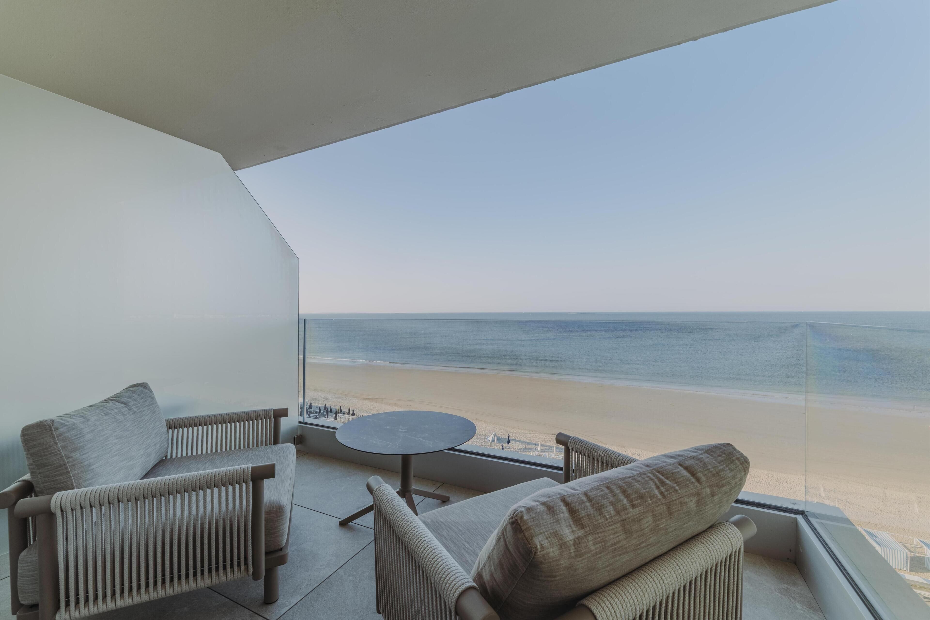 Uma varanda com vista para a praia, equipada com duas cadeiras e uma mesa pequena.