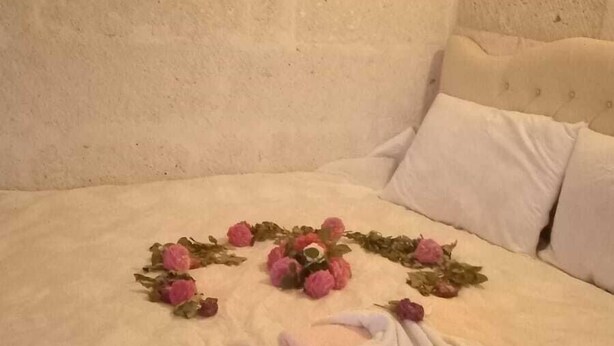 Cama com colcha branca, arranjo de flores rosa em formato de coração e folhagem verde, além de uma toalha branca.