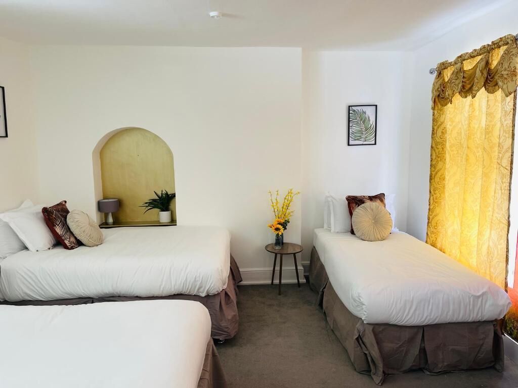 Quarto de hotel com duas camas de solteiro, uma mesa pequena com um vaso de flores, um espelho e um quadro na parede.
