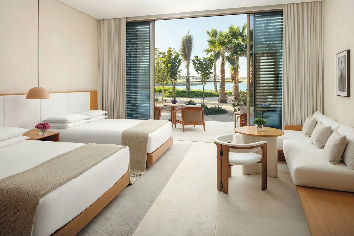 Quarto de hotel moderno com duas camas, um sofá, uma mesinha e vista para palmeiras e para a água.