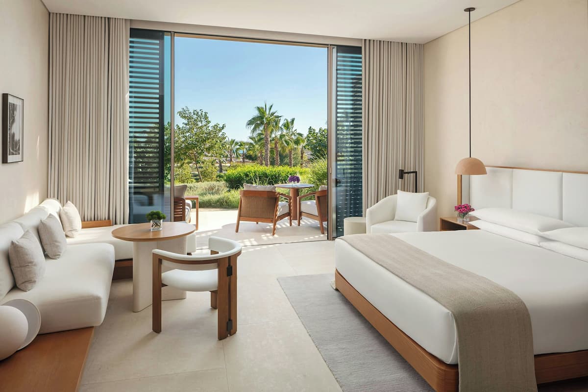 Quarto de hotel moderno com uma cama grande, um sofá, uma pequena mesa e uma cadeira. Há uma varanda com vista para palmeiras e vegetação.