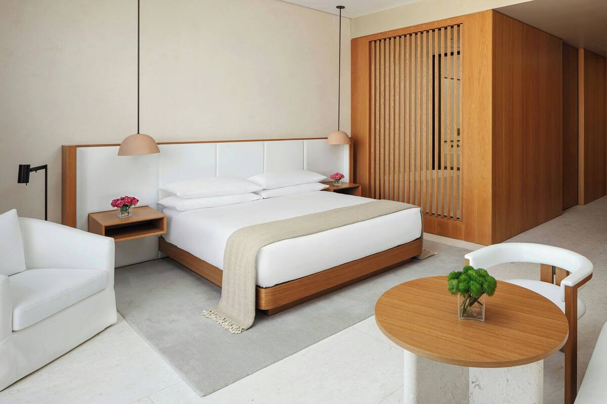 Quarto de hotel moderno com cama, mesas de cabeceira, sofá, mesa de madeira redonda e uma planta decorativa.