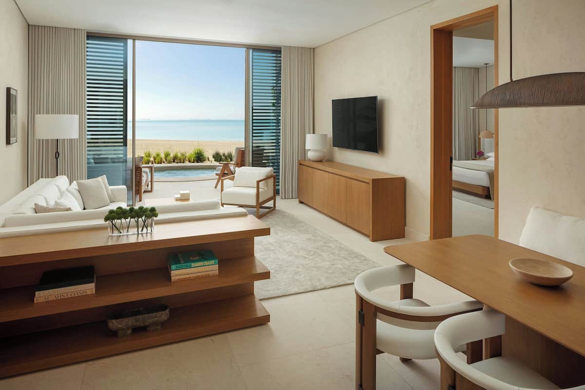 Quarto de hotel moderno com uma grande janela com vista para a praia, uma mesa de jantar de madeira, um sofá e uma TV na parede.