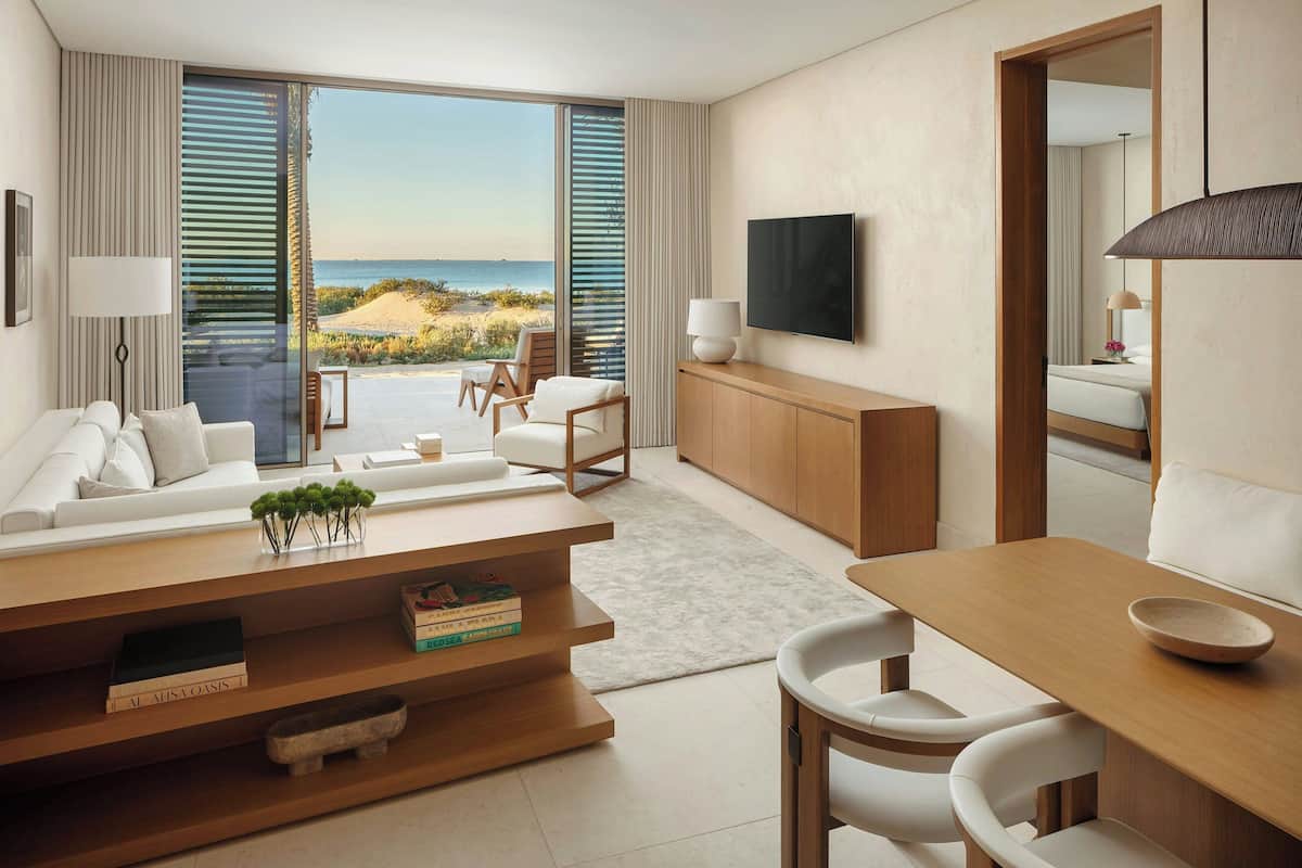 Uma sala de estar moderna com sofá, mesa de jantar, cadeiras, televisão fixada na parede e vista para a praia através de grandes janelas.