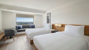 Quarto de hotel com duas camas, um sofá, uma mesa pequena e uma janela com vista para a cidade.