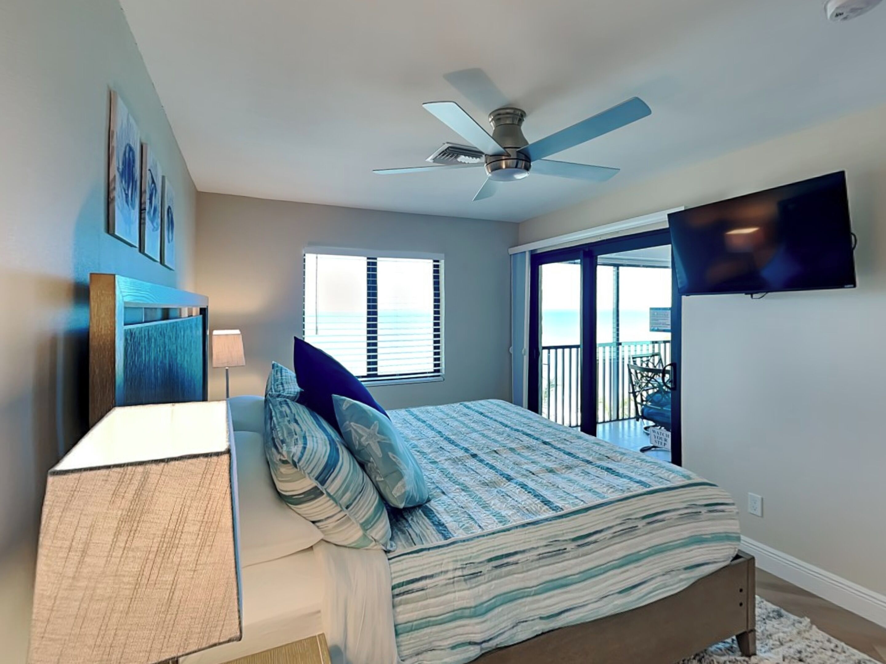Quarto com cama, ventilador de teto, TV, janela com persianas e vista para o mar.