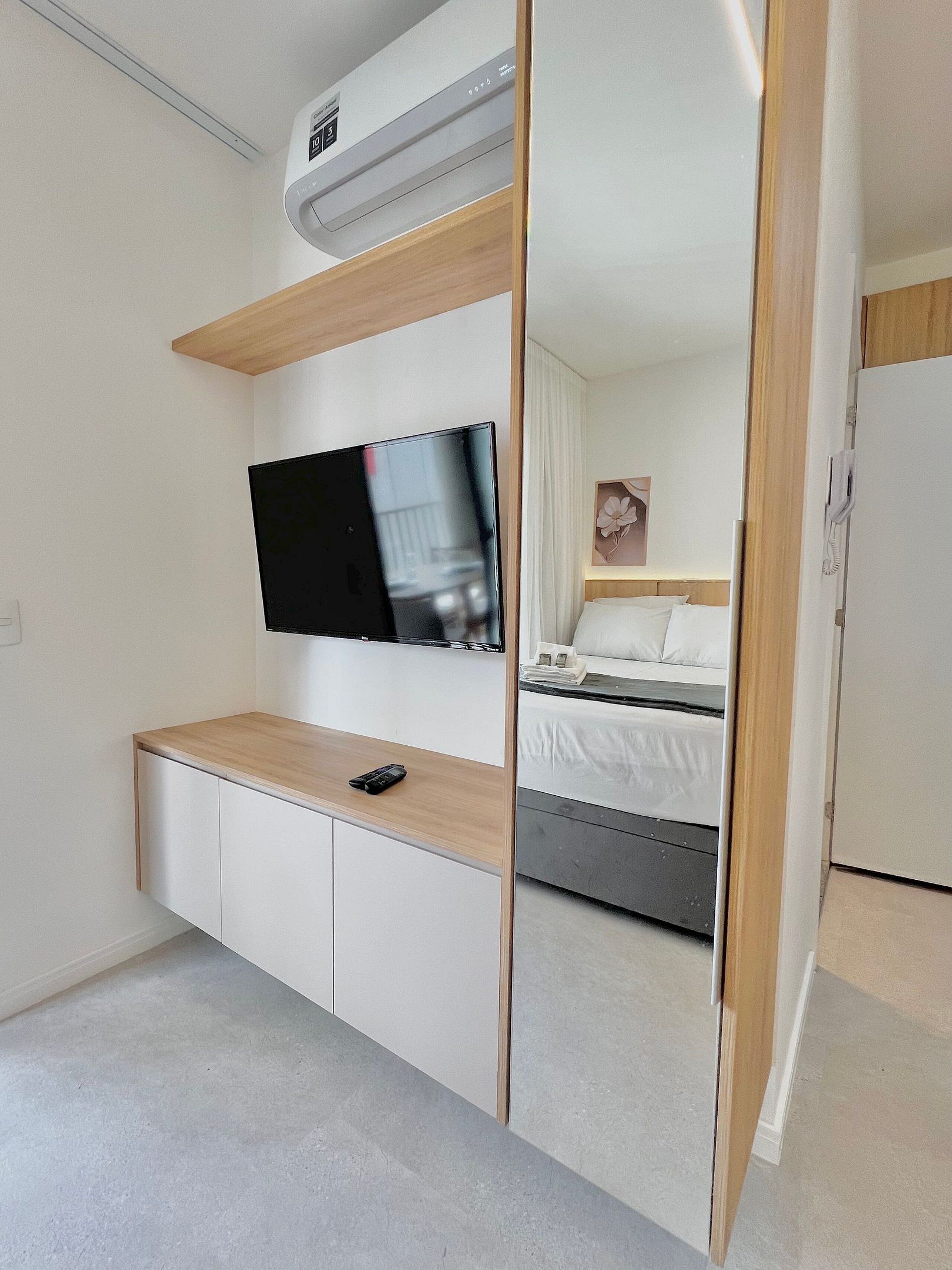 Quarto de hotel moderno com televisão de tela plana fixada em uma prateleira de madeira, mesa de cabeceira com controle remoto, cama com roupa de cama branca e um espelho refletindo o quarto.