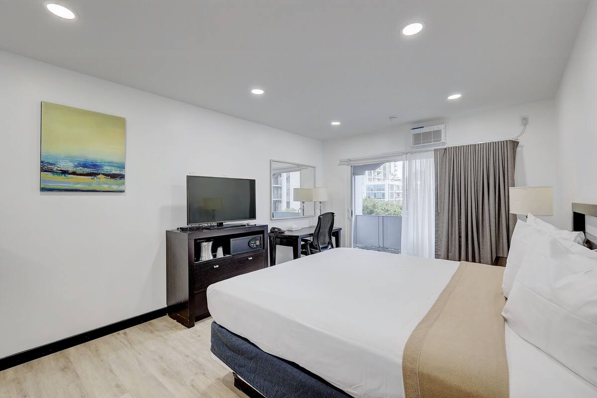 Quarto de hotel com uma cama, uma escrivaninha com computador, televisão e um quadro na parede.