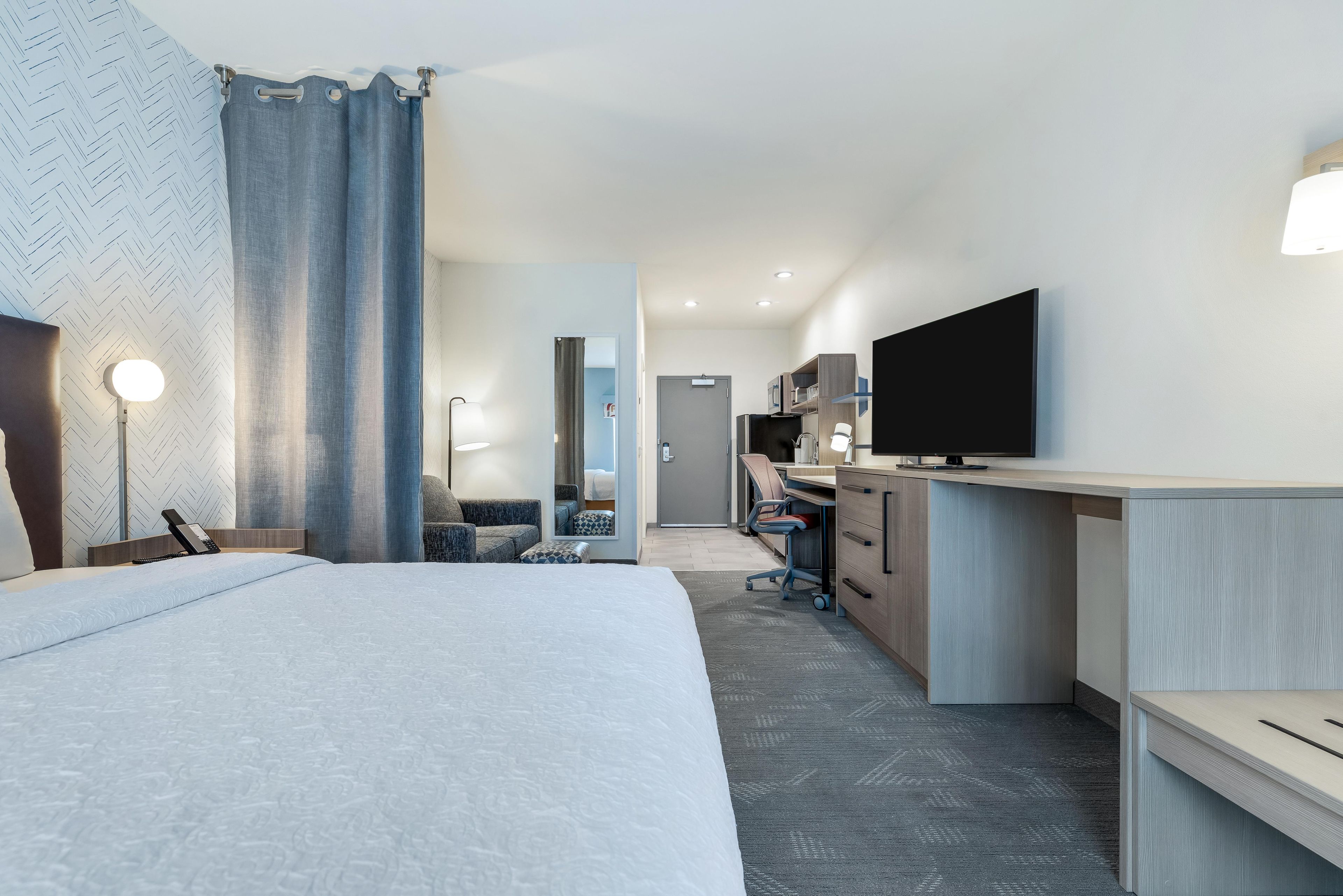 Quarto de hotel moderno com uma cama grande, uma escrivaninha com televisão de tela plana, um espelho e um banheiro visível por uma porta aberta.