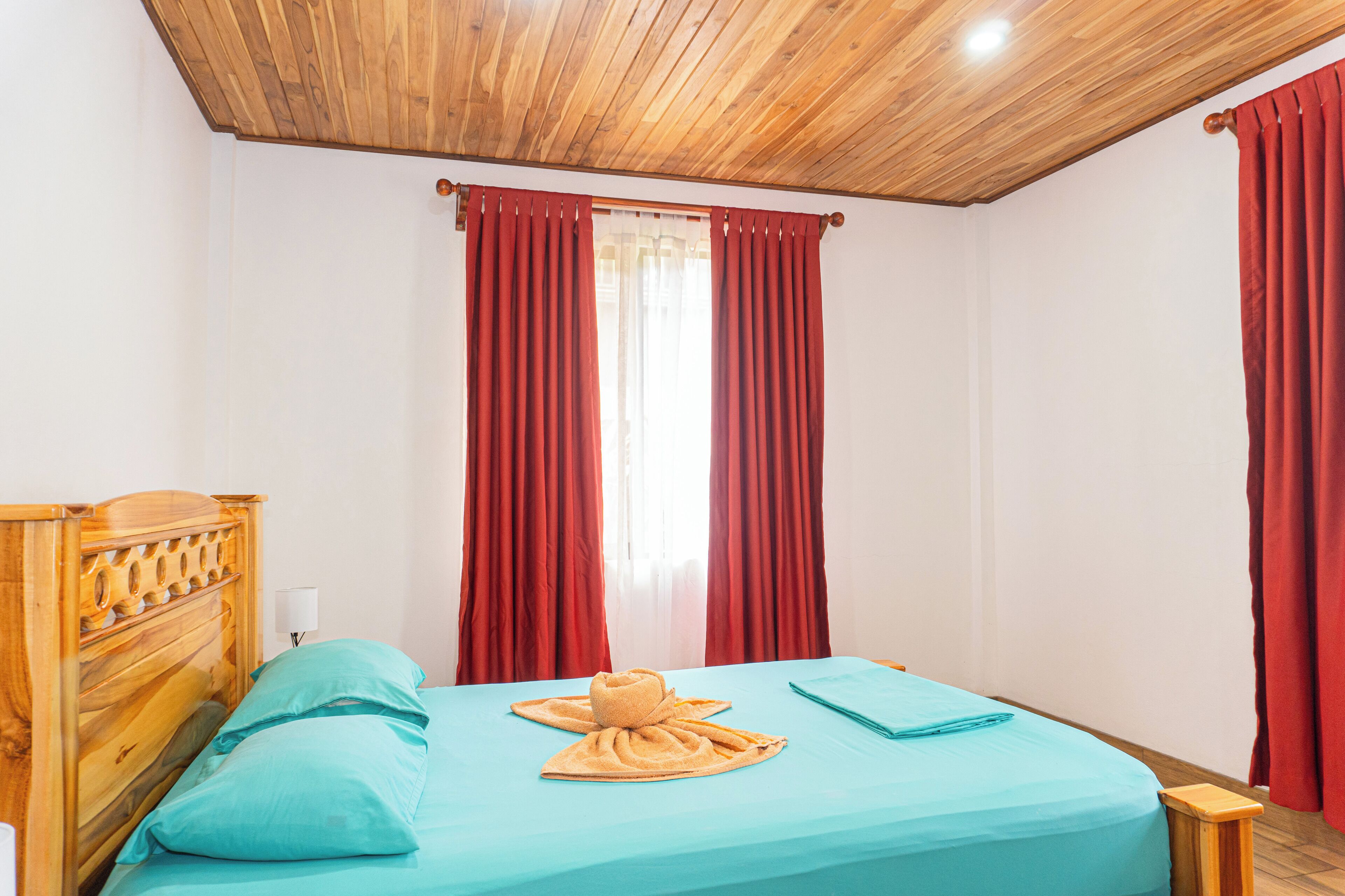 Quarto com teto de madeira, cama com colcha azul, cortinas vermelhas e cabeceira de madeira.