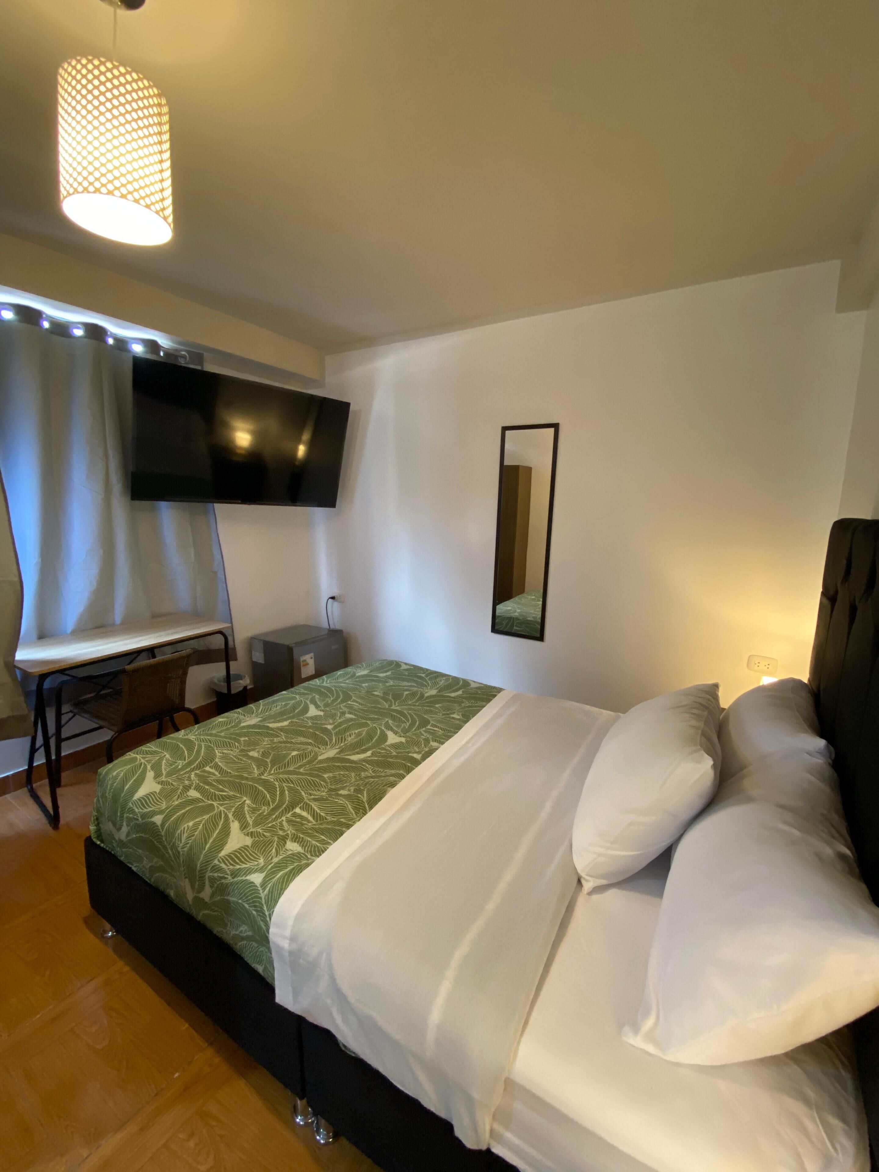 Quarto de hotel com cama, escrivaninha, cadeira, televisão e espelho.