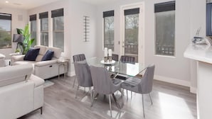 Uma sala de estar moderna com uma mesa de jantar de vidro, cadeiras cinzas, um sofá branco e uma janela grande.