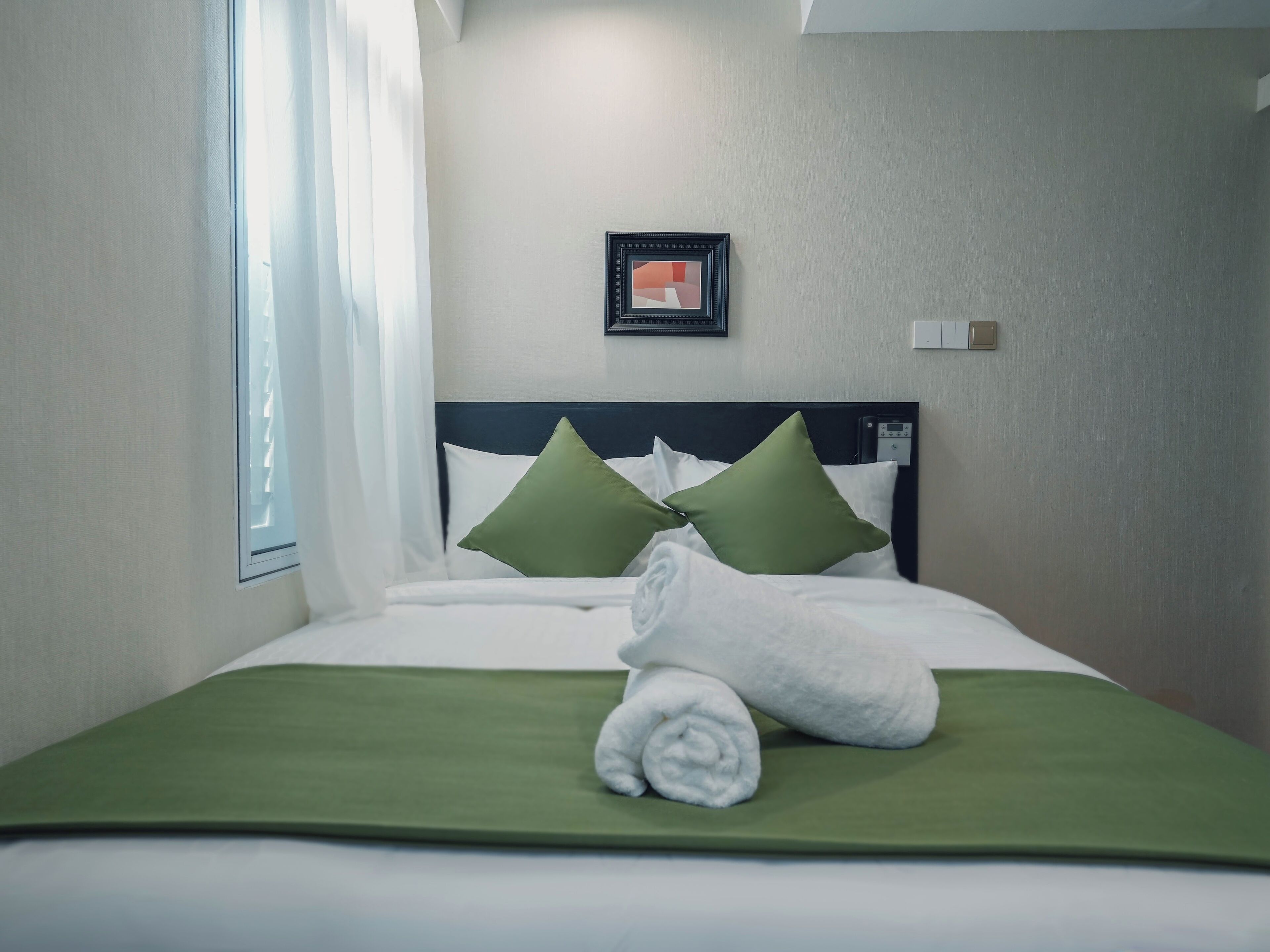 Cama bem arrumada com roupa de cama e almofadas verdes, toalha branca enrolada na cama e um quadro na parede.