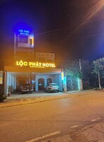 Hotel Lôc Phát Hotel, com letreiro de neon, entrada com portão e dois carros estacionados na frente.