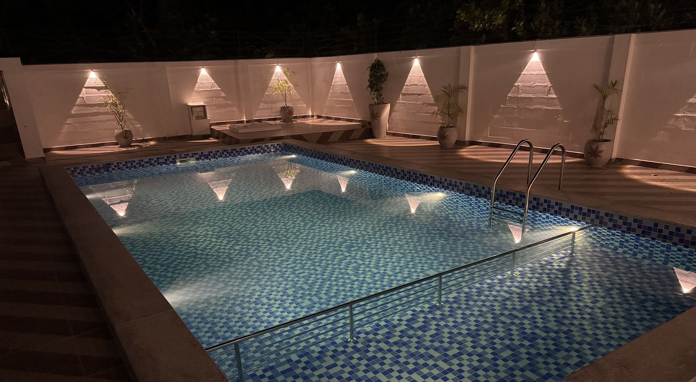 Piscina com padrão de azulejos quadriculados, rodeada por um deck de pedra e vasos de plantas.