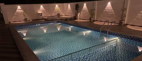 Piscina com padrão de azulejos quadriculados, rodeada por um deck de pedra e vasos de plantas.
