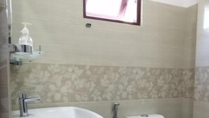 Casa de banho com lavatório branco, sanita e chuveiro. Há uma janela sobre o lavatório.
