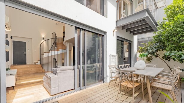 Uma casa moderna com um deck de madeira, uma área de refeições com cadeiras de vime e uma escadaria que leva a um andar superior.