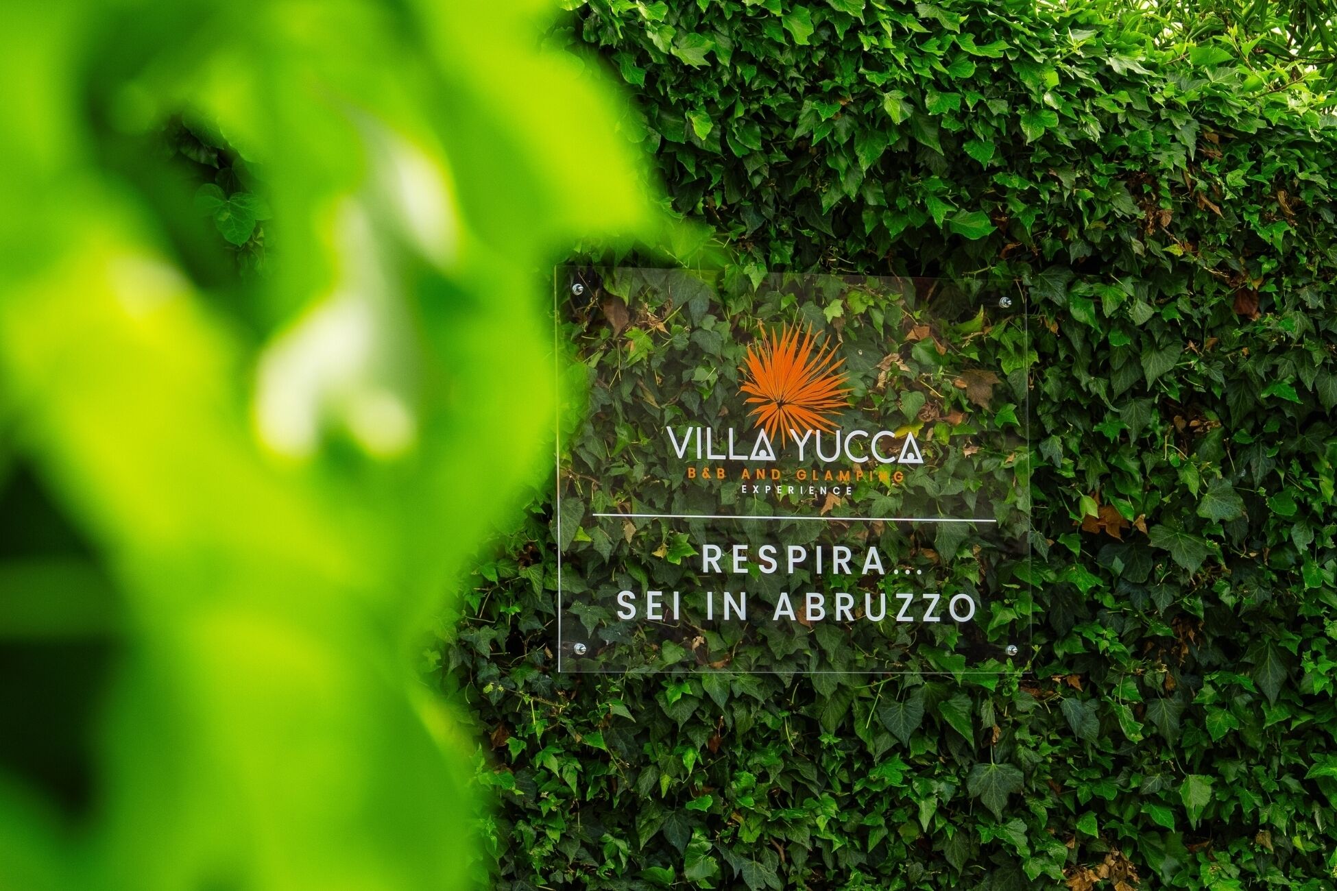 Placa com o texto 'Villa Yucca B&B and Glamping Experience' e 'Respira Sei in Abruzzo' ao fundo, folhagem verde.