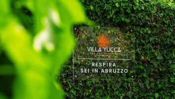 Placa com o texto 'Villa Yucca B&B and Glamping Experience' e 'Respira Sei in Abruzzo' ao fundo, folhagem verde.