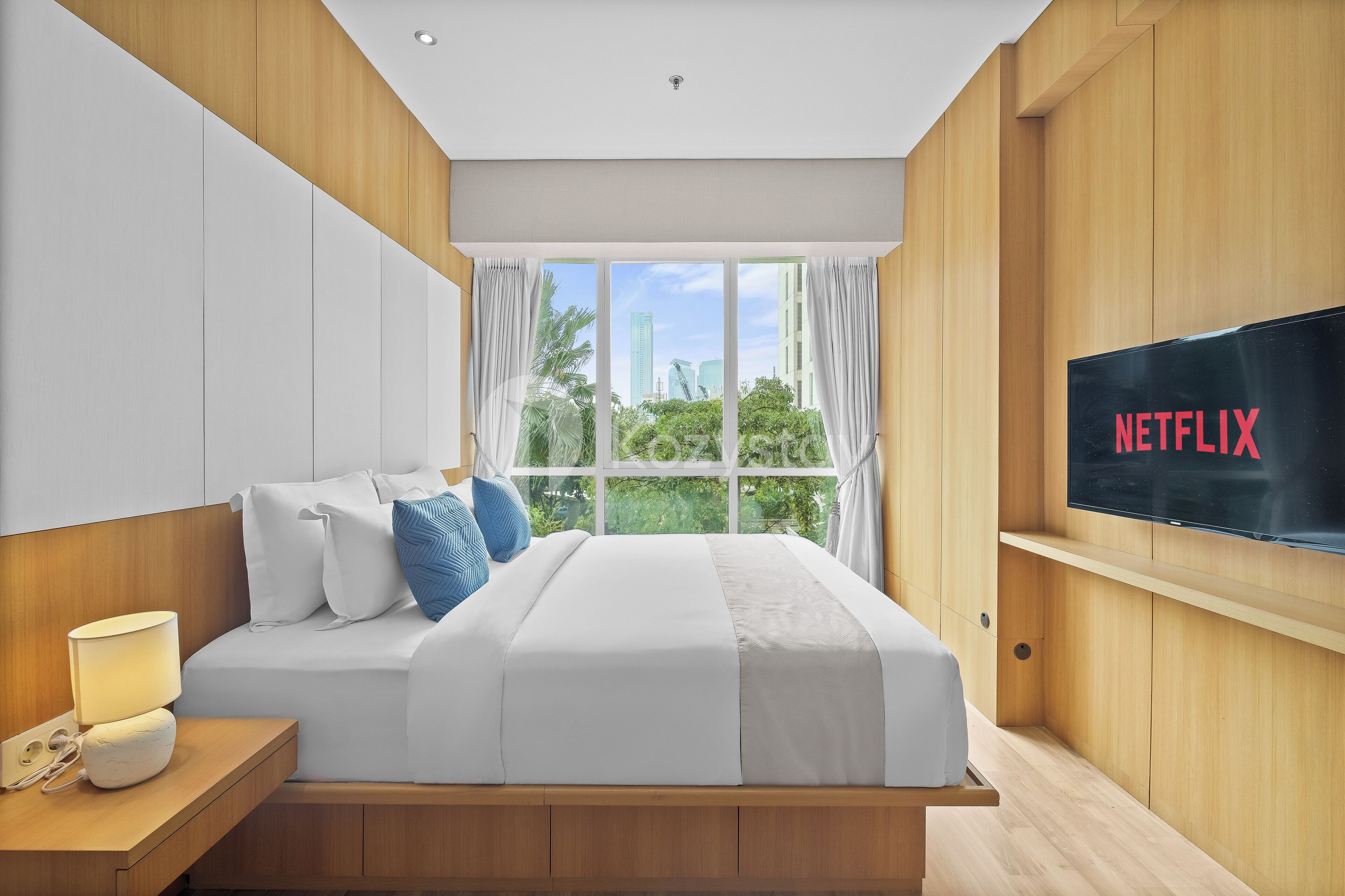 Quarto de hotel moderno com uma cama grande, uma mesa de cabeceira com abajur, televisão fixada na parede e uma janela com vista para edifícios e áreas verdes.