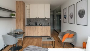 Quarto de hotel moderno com cozinha compacta, área de refeições, espaço de estar com cadeira e mesa, e obras de arte emolduradas na parede.