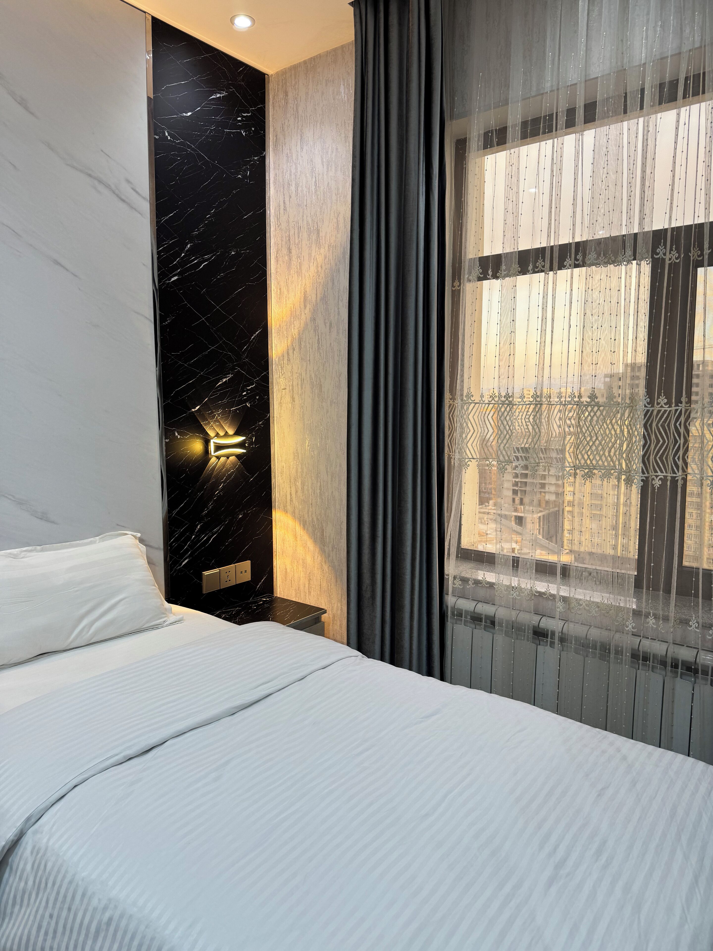 Quarto moderno com cama, parede de mármore, janela com cortinas e vista para a cidade.