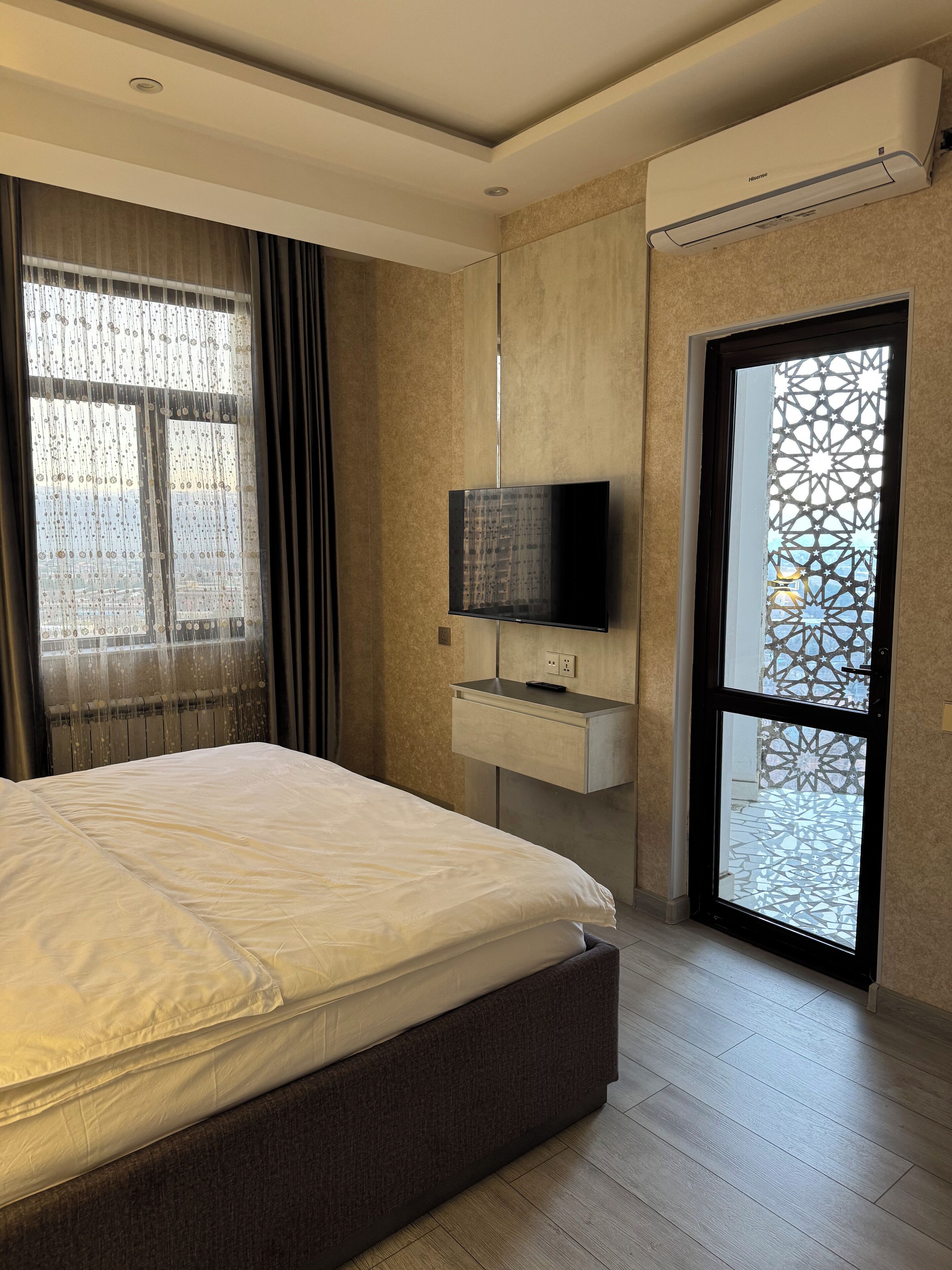 Quarto de hotel moderno com uma cama grande, televisão de tela plana na parede, ar condicionado embutido e janela decorativa com padrão geométrico.