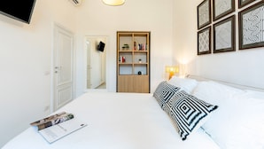 Quarto com uma cama grande, uma mesa de cabeceira com uma revista, um guarda-roupa de madeira, uma TV e arte de parede estampada.