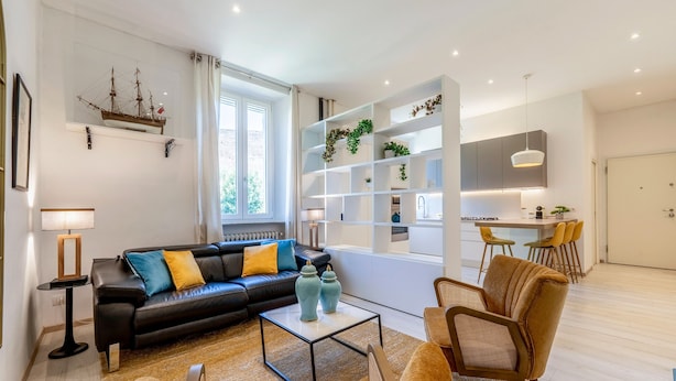Uma sala de estar moderna com um sofá preto, uma mesa de centro e uma poltrona marrom. Há uma estante com plantas e uma área de jantar com cadeiras amarelas.