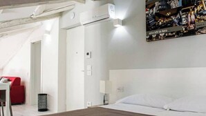 Quarto de hotel com uma cama, uma escrivaninha, uma cadeira vermelha e uma prateleira fixada na parede com objetos.