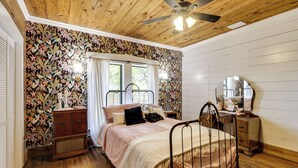 Um quarto com teto de madeira, uma cama com estrutura metálica, cômoda, espelho e papel de parede floral.