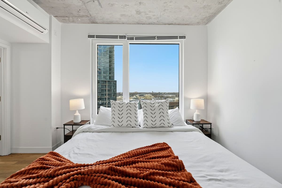 Quarto moderno com uma cama grande, duas luminárias de cabeceira e vista para o horizonte urbano através de grandes janelas.