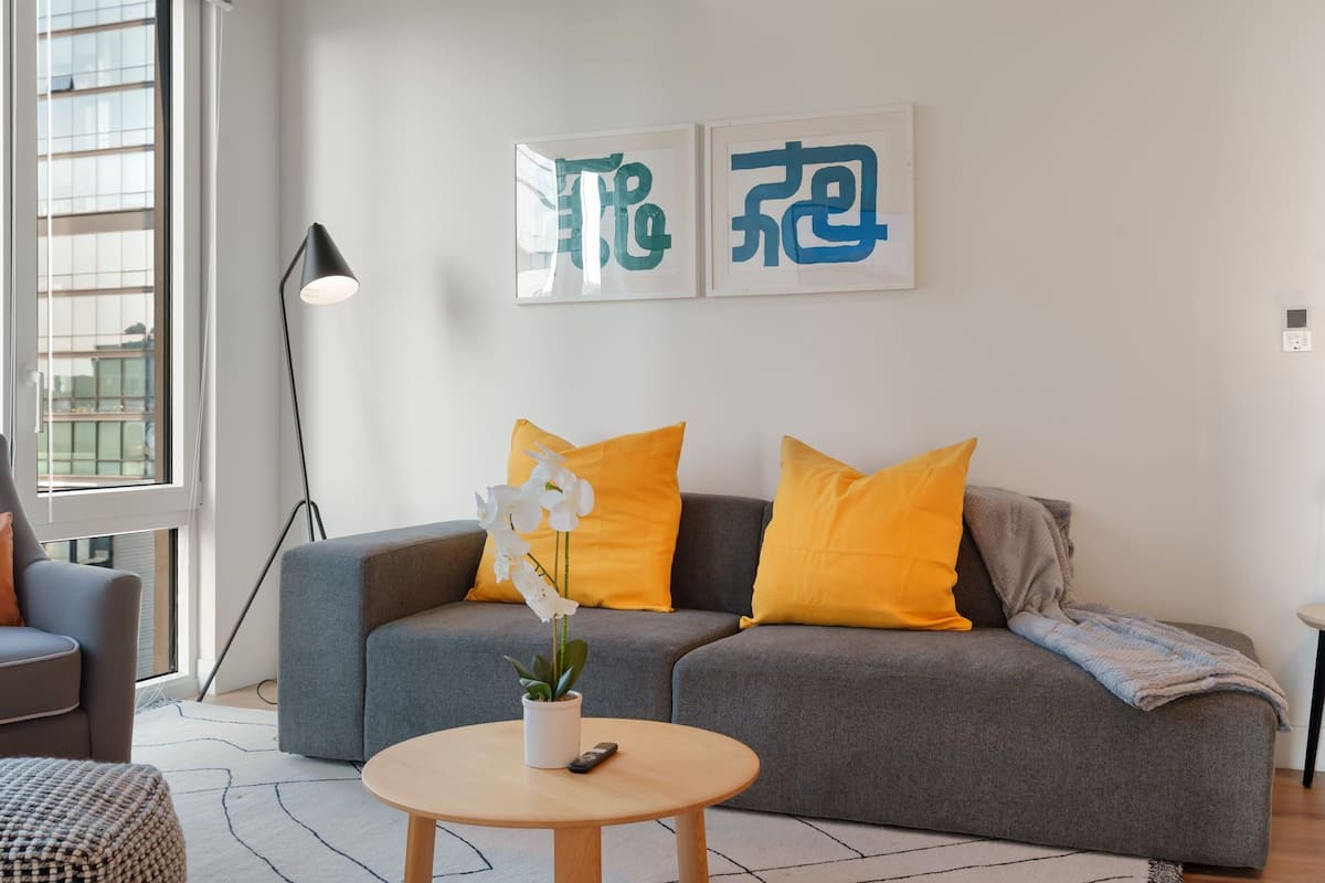 Uma sala de estar moderna com um sofá cinzento, almofadas amarelas, uma mesinha com uma planta e duas obras de arte emolduradas na parede.