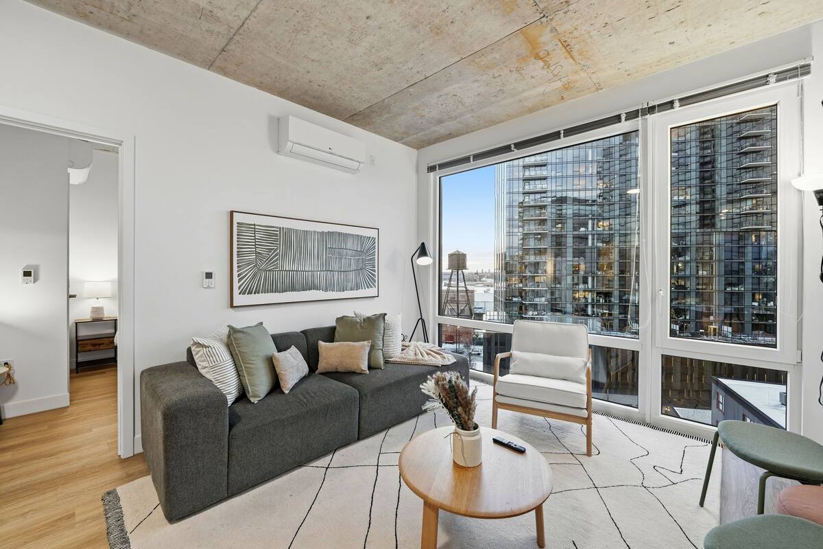 Uma sala de estar moderna com um sofá cinzento, uma poltrona branca e uma mesa de centro redonda. Uma janela grande oferece vista para a paisagem urbana.