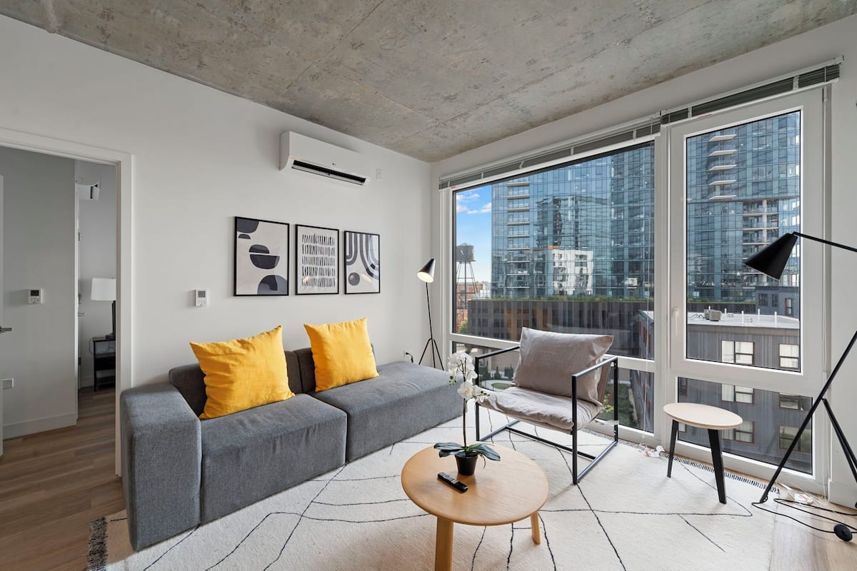 Uma sala de estar moderna com um sofá cinza, almofadas amarelas, uma mesa de centro redonda e uma grande janela com vista para a cidade.