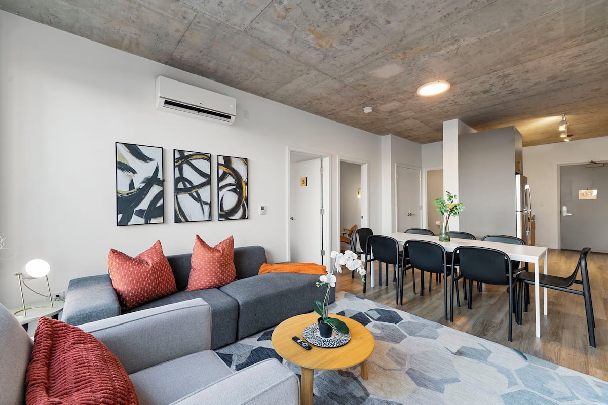 Uma sala de estar moderna com um sofá cinza, almofadas vermelhas, uma mesa de centro redonda de madeira e arte abstrata nas paredes.