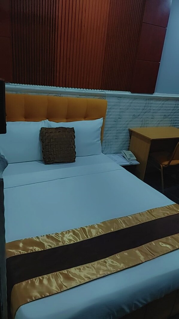 Uma cama com colcha branca e amarela, travesseiro trançado marrom e cabeceira de madeira. Há uma pequena mesa com uma cadeira ao lado da cama.