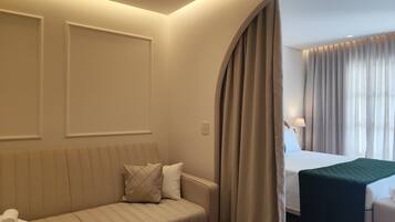 Quarto de hotel com sofá, uma pequena mesa de madeira, cama com colcha verde e janela com cortinas.