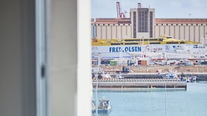 Uma varanda com vista para um porto, incluindo um navio grande com a sinalização 'FREI OLSEN' e 'Ferry', e um pequeno barco atracado nas proximidades.