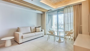 Quarto de hotel moderno com sofá, mesa de jantar com cadeiras e vista para a cidade por grandes janelas.